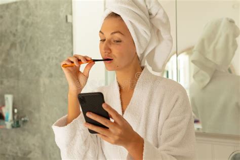 Happy Caucasian Woman Wearing Robe Brushing Teeth And Using Smartphone