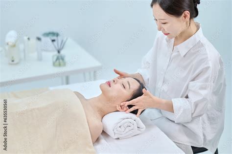 아시아 한국의 젊은 여성이 스파 또는 피부관리전문점 또는 피부과 병원에서 침대에 누워 베이지색 대형 타올을 덮고 눈을 감고있고 하얀색 셔츠를 입은 전문관리사가 손으로 얼굴