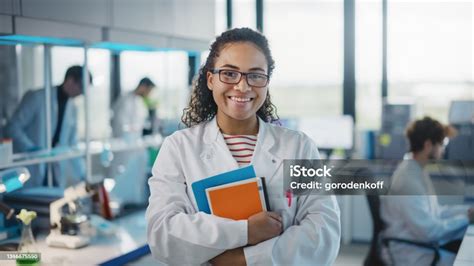 의학 연구소 흰색 코트와 안경을 착용 아름다운 똑똑한 젊은 흑인 과학자 테스트 책을 보유하고 카메라를보고 미소 다양한 전문가 팀 중간 초상화 샷 학생에 대한 스톡 사진 및