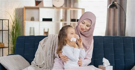 사랑스러운 돌보는 무슬림 엄마 집에서 파란색 소파에 앉아 무릎에 어린 딸을 안고 있습니다 귀여운 행복한 아이는 위생 절차를