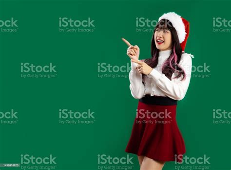 흰색과 빨간색 크리스마스 테마 의상을 입은 젊은 예쁜 아시아 여성이 산타 모자 손가락을 입고 복사 공간 녹색 화면 배경을