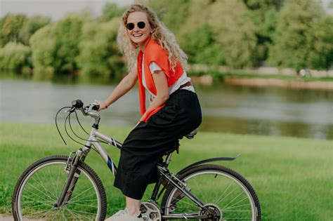 쾌적한 찾고 만족 곱슬 머리 여자는 야외에서 자유 시간을 보내고 자전거를 타고 혼자 여가를 보내고 자연의 신선함을 즐기고 즐거운 미소로 카메라를 봅니다 여름 시간입니다 25
