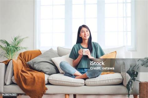 집에서 소파에서 휴식을 취하며 커피를 마시는 행복한 젊은 아시아 여성 현대 거실의 소파에 앉아 휴식을 즐기는 미소 짓는 여성