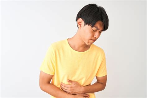노란 캐주얼 티셔츠를 입은 중국인 남성은 메스꺼움 고통스러운 질병이 좋지 않기 때문에 위장에 손을 얹고 고립 된 흰색 배경 위에 서 있습니다 아쉬 컨셉 T 셔츠에 대한 스톡