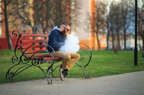 Vape입니다 큰 수염과 선글라스에서 유행 헤어스타일 젊은이 잔인 한 도시 공원에 빨간 벤치에 전자 담배 연기가 난다 스팀 클라우드입니다 공원에 대한 스톡 사진 및 기타
