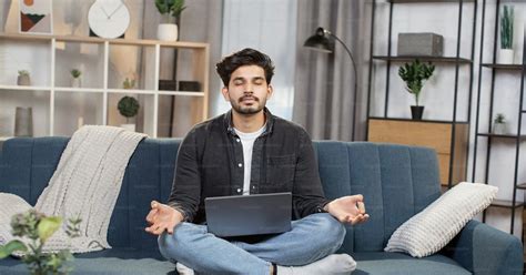 유쾌하고 호감이 가는 젊은 아라비아 인도 남자는 거실의 소파에 노트북 Pc를 들고 연꽃에 앉아 눈을 감고 미소를 지으며 명상을 하고 무드라에서 손가락을 접습니다 사진