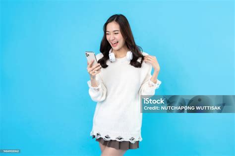 긴 머리 캐주얼 웨어 흰색 무선 헤드폰을 착용한 아름다운 젊은 백인 아시아 여성 회색 스마트 폰을 사용하여 흥분하고 재미있게 지냅니다 스튜디오의 파란색 배경에 25 29세에