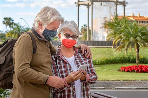 코로나바이러스로 인해 외과 용 마스크를 쓰고 흰색 머리와 배낭 여행자를 가진 선임 부부는 도시 투어 중에 휴대 전화를 봅니다 여행과 자유를 즐기는 활동적인 은퇴자 2명에
