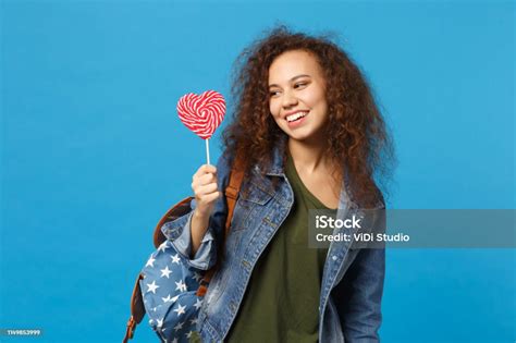 데님 옷 배낭은 파란색 벽 배경 스튜디오 초상화에 고립 된 사탕을 잡아 젊은 아프리카계 미국인 소녀 십 대 학생 고등학교 대학 대학 개념에서 교육 복사 공간을 모의 가방에