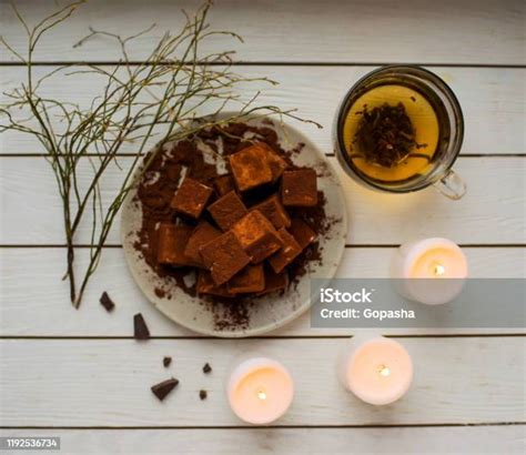 코코아 가루로 덮인 수제 수플레 큐브는 밝은 나무 테이블 위에 접시에 놓여 있고 유리 잔에 뜨거운 차와 그 옆에 는 세 개의 흰색 불타는 양초 초콜릿 조각 및 잔가지가