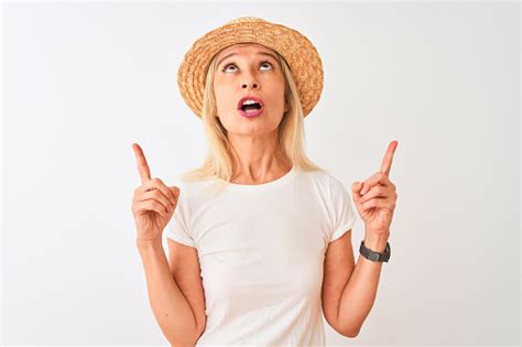 캐주얼 한 티셔츠와 모자를 쓰고 고립 된 흰색 배경 위에 서있는 중년 여성은 손가락과 팔을 들고 위를 올려다 보고 놀랐습니다 T