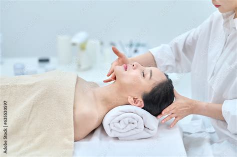 Foto Stock 아시아 한국의 젊은 여성이 스파 또는 피부관리전문점 또는 피부과 병원에서 침대에 누워 베이지색 대형 타올을 덮고 눈을 감고있고 하얀색 셔츠를 입은 전문