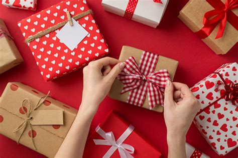 발렌타인 데이 장식 선물과 고립 된 빨간 배경에 공예 종이 선물 상자에 체크 장식 리본 활을 묶는 젊은 여성의 손의 1 인칭 탑 뷰 사진 갈색 용지에 대한 스톡 사진 및