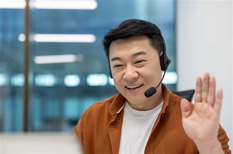 원격 통신을 위해 화상 통화를 사용하는 아시아 남성 사업가 현대적인 사무실 환경에서 헤드셋 착용 온라인 회의 중 미소를 지으며 손을 흔들며 기술을 선보이는 모습 경영자에