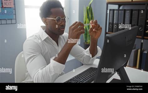 Young Man Working In A Modern Office Focusing Intently On A Pen While Seated At A Computer Desk