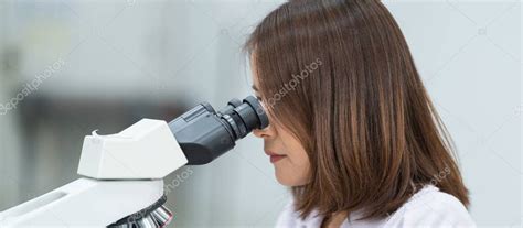 Молодая ученая женщина в лабораторном халате смотрит в микроскоп в лаборатории для проведения