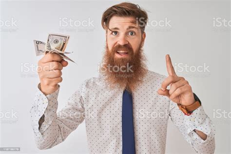 젊은 빨간 머리 아일랜드어 사업가 는 행복한 얼굴 번호 하나 와 손가락을 가리키는 아이디어 또는 질문으로 놀란 고립 된 흰색 배경 위에 서 있는 달러를 들고 가리키기에 대한