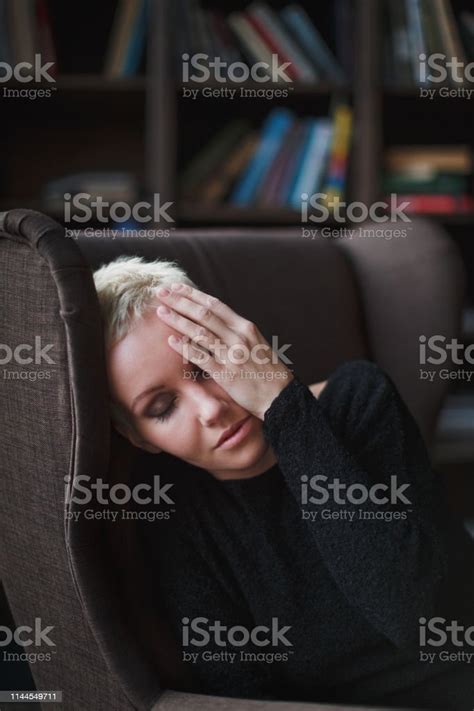 금발 짧은 머리를 가진 아름 다운 부드러운 젊은 여성은 어두운 벽 방에 있는 안락의 자에 앉아 한 손으로 그녀의 얼굴을 덮고 배경에 책이 있는 책장이 있습니다 수직 사진