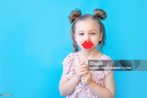 파란색 고립된 스튜디오 배경에 있는 어린 소녀가 입술이 있는 막대기에 종이 액세서리를 들고 재미있게 놀고 있습니다 텍스트 복사를 위한 공간입니다 귀여운 아이 6 7 살에