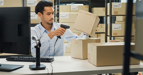 행복한 아시아 남자는 공식적인 유니폼 셔츠를 입고 책상 앞에 앉아 컴퓨터와 바코드 기계 스캐너를 사용하여 창고에서 골판지 상자