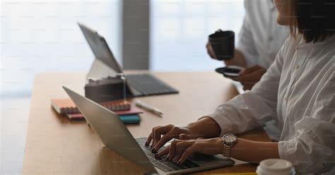 아름다운 여성이 컴퓨터 태블릿에 타이핑하는 동안 질서 정연한 거실 위의 나무 작업 테이블에서 동료 옆에 배경으로 앉아 있는 자른 이미지 집에서 일하는 개념 사진