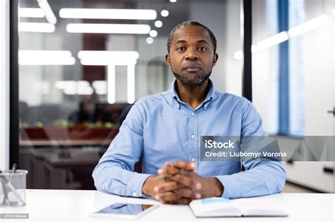 현대적인 사무실의 책상에 앉아 두 손을 깍지 낀 자신감 넘치는 사업가는 결단력과 집중력을 반영합니다 책상 위에 펼쳐진 책과 스마트폰이 있는 전문적인 분위기 남자에 대한 스톡