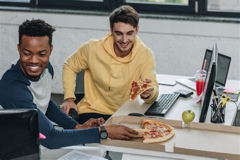Premium Photo Two Cheerful Multicultural Programmers Eating Pizza While Sitting At Desks In Office