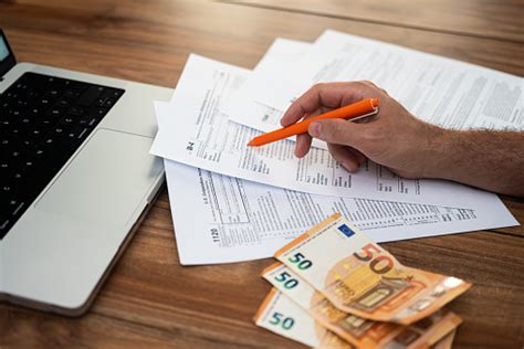 노트북과 유로 지폐 옆의 나무 책상에서 손으로 세금 양식을 작성하고 있습니다 세금 준비 및 재정 문서화가 진행 중입니다 회계 재무 계획 및 세금 신고 기간 Internal