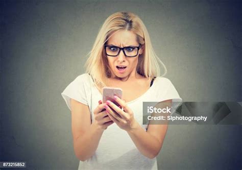 충격적인 메시지입니다 휴대 전화를 들고와 회색 배경에 서 있는 동안 그것을 보고 놀된 여자 여자에 대한 스톡 사진 및 기타