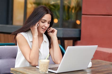 검은 긴 머리와 매력적인 여자의 총 비즈니스 파트너와 함께 휴대에 서핑 인터넷과 거리 작업용 노트북 컴퓨터를 사용 하 여 칵테일 음료 야외 아늑한 카페에서 포즈 노트북에