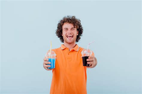 카메라에 곱슬 머리 미소와 잘 생긴 소년 오렌지 셔츠 고립 된 파란색 배경 긍정적 인 얼굴 감정 복사 공간을 입고 주스와 두 개의 플라스틱 컵을 보유 곱슬 머리에 대한 스톡