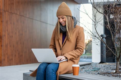 벤치에 앉아 활짝 웃는 젊은 여자는 도시 거리에서 커피를 손에 들고 노트북을 본다 사무실이 아닌 관리자는 랩톱에서 추가 작업에 종사하고 있습니다 거리에 대한 스톡 사진 및