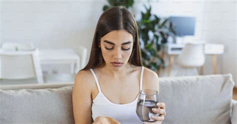 젊은 여자는 손에 물 한 잔과 함께 알약을 복용합니다 스트레스를 받은 여성은 창문을 들여다보고 진정제가 함유된 항우울제를 마신다 여자는 우울함을 느끼고 마약을 복용합니다