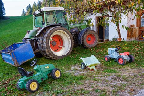 어린이용 페달 트랙터 두 대와 트레일러 한 대가 빨간 바퀴가 달린 초록색 트랙터와 트레일러 앞에 주차되어 있습니다 배경에는 농가와 초원이 있습니다 구릉 풍경 0명에 대한