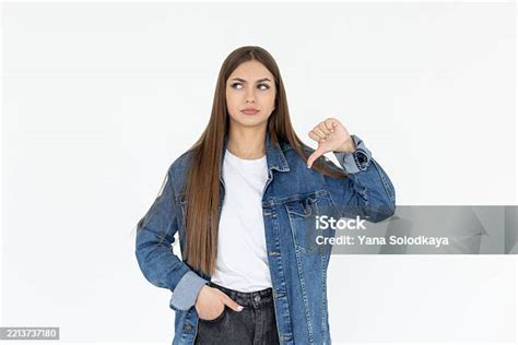 젊은 슬프고 불쾌한 불행한 여자 흰색 티셔츠와 청바지 코트를 착용하고 엄지 손가락을 아래로 보여주는 싫어하는 제스처 흰색 배경 스튜디오 초상화에 고립되어 있습니다 사람들