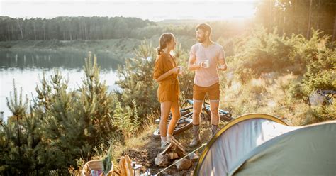 젊은 부부는 캠프장에 서서 일몰에 호수 근처의 산을 여행하는 동안 뜨거운 음료를 즐기고 이야기합니다 사진 Unsplash의 우크라이나 이미지