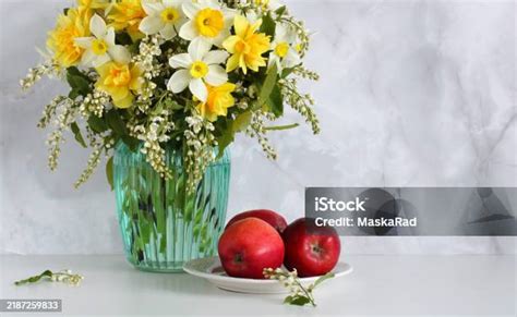 밝은 노란색 수선화와 흰 새 벚꽃이 접시에 신선한 빨간 사과와 함께 꽃병에 배열되어 대리석 표면에 생생한 봄 장식을 만듭니다