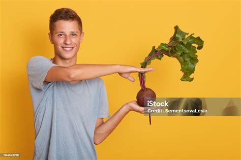 노란색 배경 위에 고립 된 잘 생긴 모델의 실내 스튜디오 샷 한 손에 생 야채를 들고 크기를 보여주고 제스처를 하고 진심으로 미소 사람과 건강한 음식 개념 갈색 머리에 대한