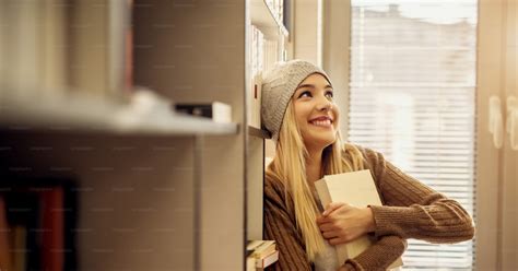 교육 도서관 학생 및 사랑의 개념 사랑에 빠진 행복한 힙스터 고등학생 소녀는 도서관의 책장에 기대어 책을 들고 웃고 있다