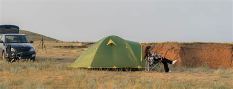 여자는 초록색 텐트 앞 캠프 의자에 앉아 있고 근처에 차가 주차되어 있다 장면은 언덕을 배경으로 풀이 무성한 들판을 배경으로 합니다 25 29세에 대한 스톡 사진 및 기타