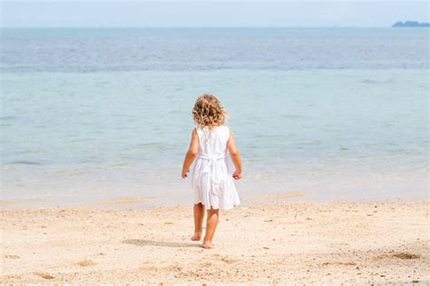 하얀 드레스를 입은 곱슬 머리를 가진 어린 소녀가 모래 사장을 걷습니다 한 아이가 해변을 따라 걷는다 아이들과 함께하는 여행 및