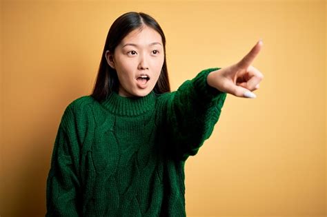 젊은 아름다운 아시아 여자 착용 녹색 겨울 스웨터 위에 노란색 고립 된 배경 가리키는 손가락 으로 앞서 놀라게 열린 입 깜짝