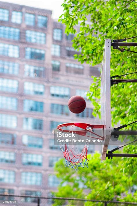 고층 빌딩을 배경으로 무성한 녹지가 있는 도심 야외 코트에서 농구를 향해 비행하는 모습은 활기찬 도시 분위기 속에서 에너지와 움직임을 표현합니다 0명에 대한 스톡 사진 및