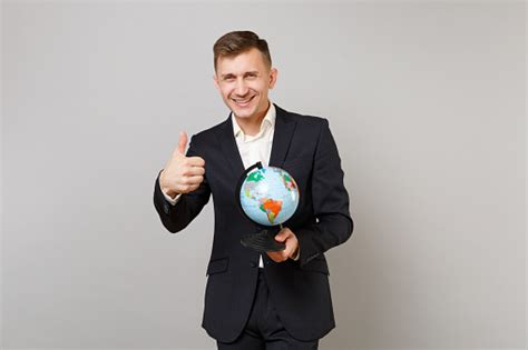 고전적인 검은 양복에 젊은 사업가를 웃 고 위로 엄지손가락을 보여주는 셔츠 회색 벽 배경에 고립 된 지구 세계 글로브를 들고 업적 경력 자산 사업 개념 복사 공간을 모의