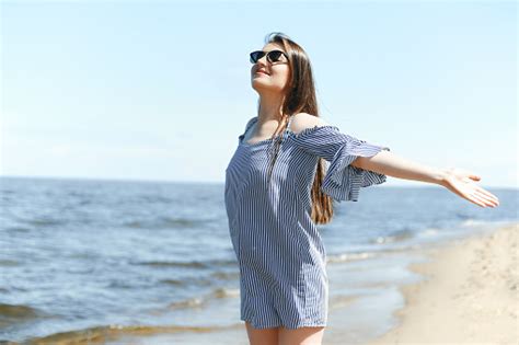 두 손을 벌리고 서 있는 바다 해변에서 자유로운 행복을 누리고 있는 행복한 미소 짓는 여자 여름 드레스를 입은 갈색 머리 여성 모델의 초상화는 야외 휴가 휴가 동안 자연을