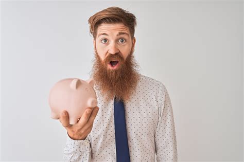 젊은 빨간 머리 아일랜드어 사업가 고립 된 흰색 배경 위에 돼지 저금통을 들고 깜짝 얼굴 두려움과 두려움 표정으로 흥분 충격에 무서워 감정에 대한 스톡 사진 및 기타 이미지