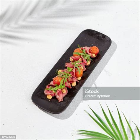 검은 접시에 체리 토마토 Ruccola 매운 소스를 곁들인 소고기를 구워냅니다 흰색 콘크리트 배경에 슬라이스 구운 쇠고기 하드 섀도우와 야자수 잎여름 스타일의 이탈리아 안티