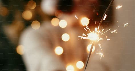 어두운 방에서 현대 크리스마스 트리 조명을 배경으로 산타 모자를 쓴 세련된 소녀의 손에 큰 폭죽이 불타고 있습니다 불꽃놀이를