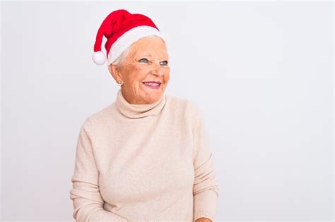 Crhistmas 산타 모자를 쓰고 고립된 흰색 배경 위에 회색 머리의 선임 여성이 얼굴에 미소를 지으며 옆으로 바라보고 자연스러운 표정 자신감 있게 웃고 있습니다 12월에