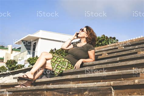 여름 화창한 날에 나무 계단에 앉아 스마트 폰으로 말하는 아름다운 소녀 이국적인 휴가를 즐기는 트렌디 한 여성 관광객 행복한 여자는 일광욕을 하고 스마트 폰과 의사 소통을